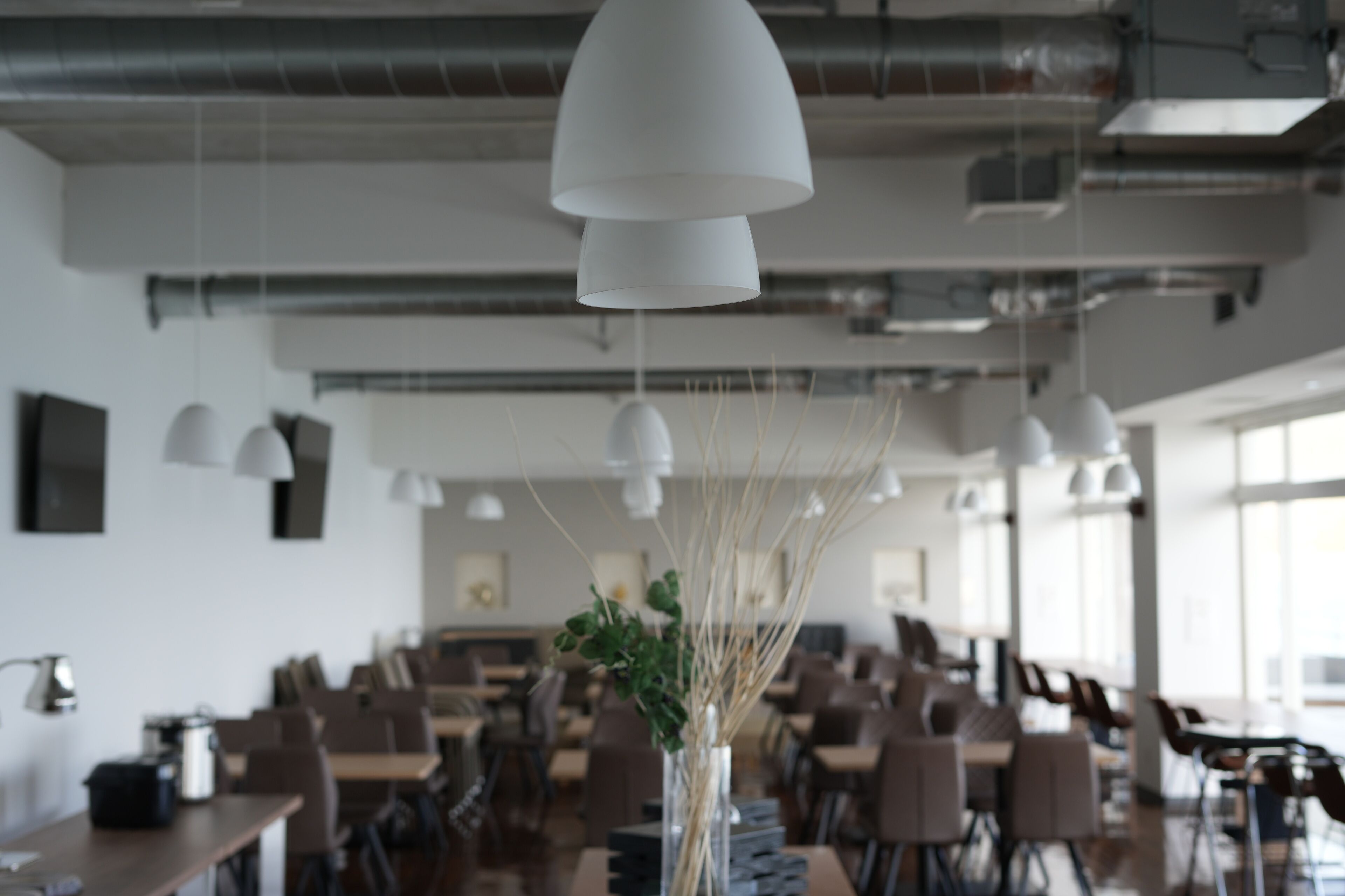 Uma sala de jantar moderna com várias mesas e cadeiras, luminárias pendentes e uma grande janela que oferece luz natural.
