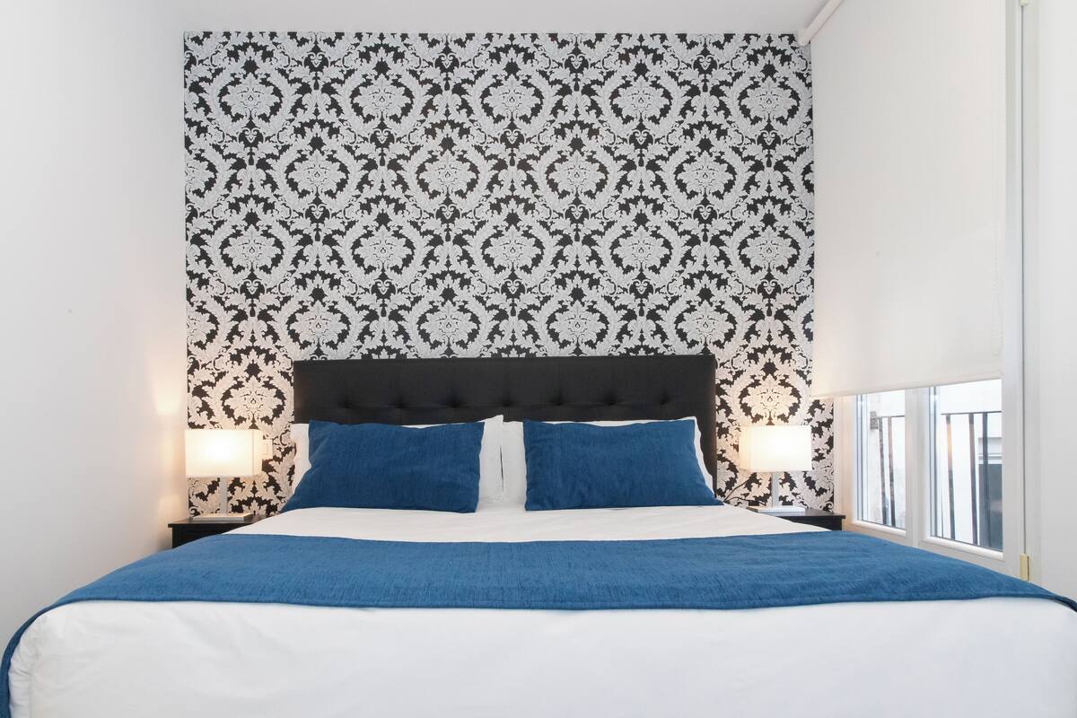 Quarto com papel de parede estampado, cabeceira estofada, duas almofadas azuis e um edredom branco.
