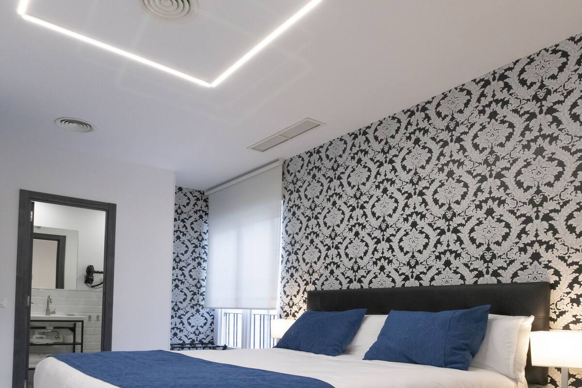 Quarto de hotel moderno com uma cama grande, uma mesa de cabeceira, um espelho e papel de parede estampado.
