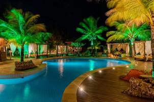 Uma área de piscina à noite, com piscina em curva, palmeiras e espreguiçadeiras.