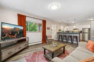 Uma sala de estar moderna com televisão, sofá e mesa de centro, cozinha com banquetas altas e uma janela grande com cortinas.