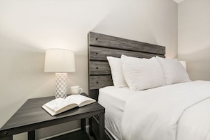 Cama bem arrumada com roupa de cama branca, mesa de cabeceira com abajur e um livro, e prateleira na parede com uma xícara.