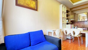 Uma sala de estar com um sofá azul, cadeiras brancas, uma mesa pequena e uma área de cozinha com revestimento de azulejo e armários de madeira.
