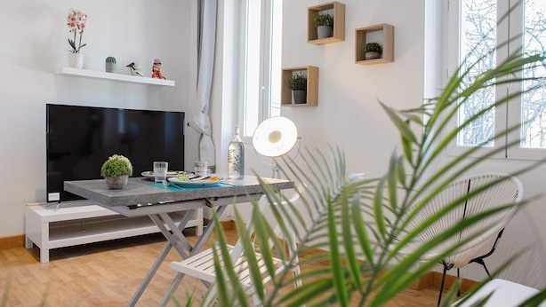 Uma sala de estar moderna com uma televisão de tela plana, uma mesa de jantar com cadeiras, um abajur de chão e diversos objetos decorativos nas estantes.