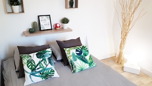 Um quarto com cama, duas almofadas estampadas com folhas tropicais, prateleira de madeira com plantas e um letreiro emoldurado, além de um vaso alto com galhos.