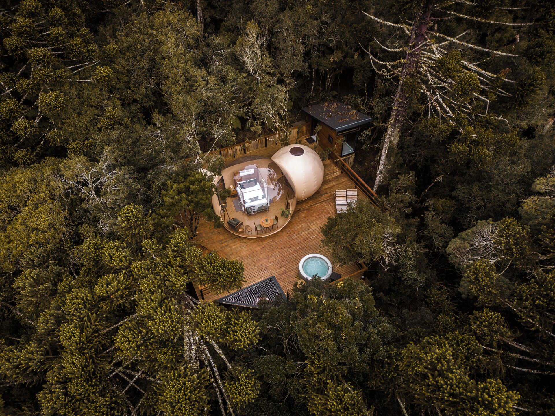 Vista aérea de uma cabana isolada, rodeada por uma floresta densa, com um deck circular, móveis de exterior, uma banheira de hidromassagem e uma pequena estrutura de madeira.