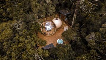 Vista aérea de uma cabana isolada, rodeada por uma floresta densa, com um deck circular, móveis de exterior, uma banheira de hidromassagem e uma pequena estrutura de madeira.