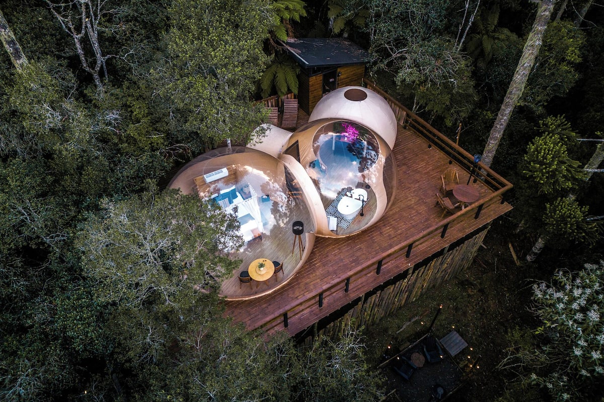 Uma casa na árvore com vários cômodos circulares, cada um com uma pequena mesa e cadeiras, cercada por uma floresta densa.