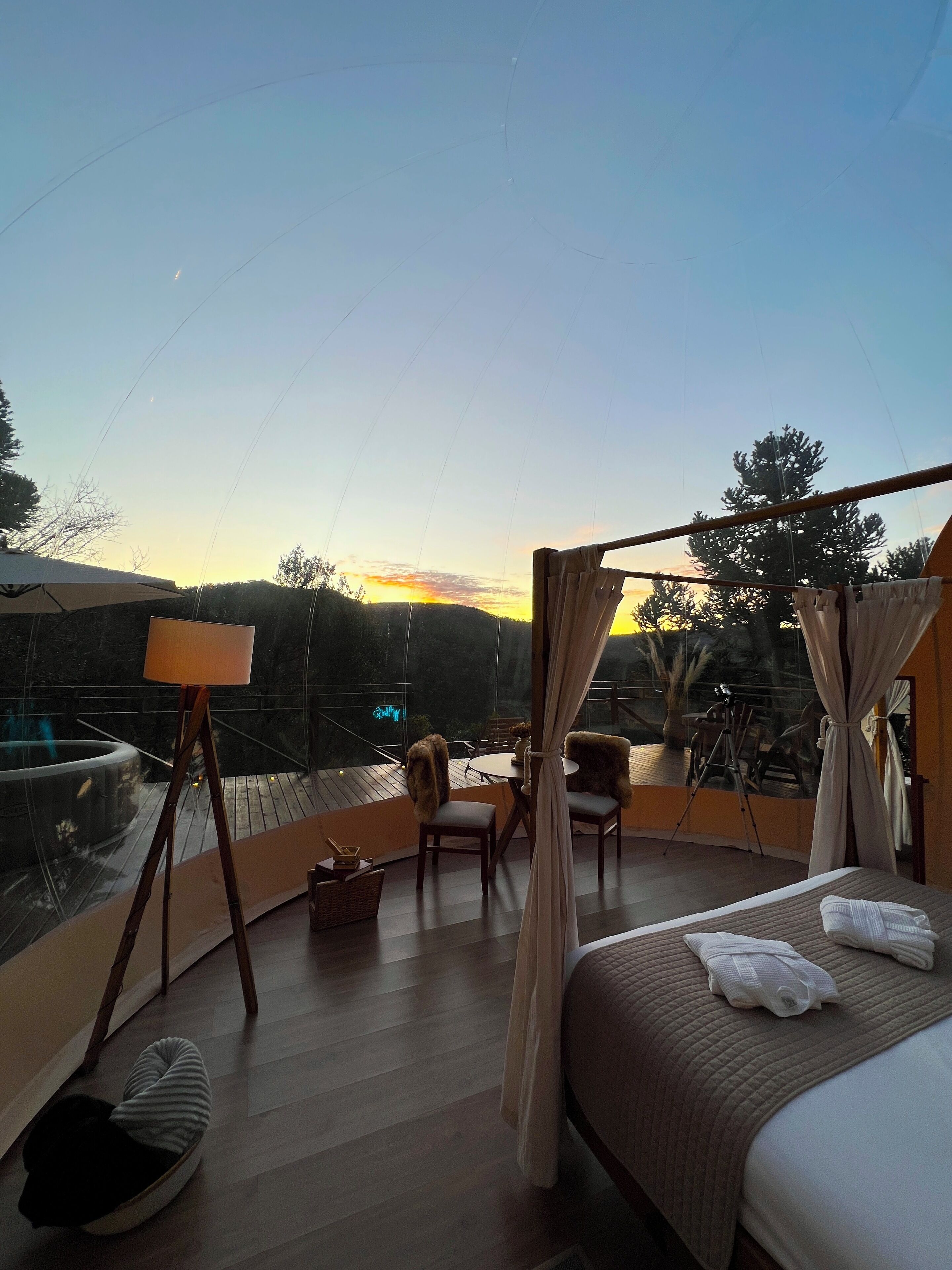 Quarto com cama, mesa de cabeceira, luminária, cadeira e vista para o pôr do sol.