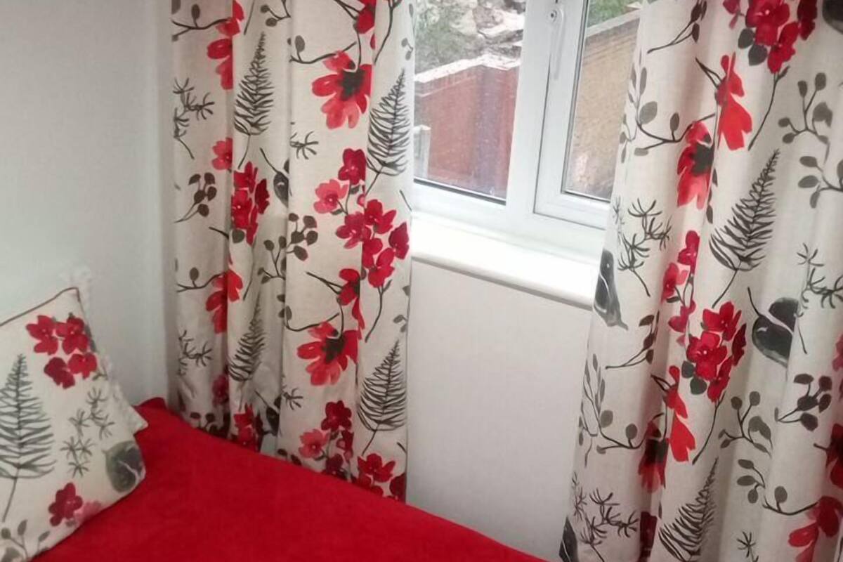Quarto com colcha vermelha, cortinas florais e janela com vista para o verde.