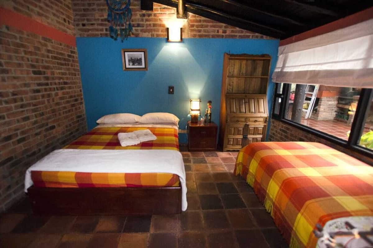 Quarto com duas camas, parede de tijolos, parede azul de destaque, armário de madeira e janela com vista.