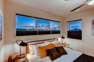 Quarto de hotel com cama, mesa de cabeceira, luminária e vista para a cidade através de grandes janelas.