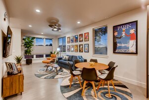 Uma sala de estar moderna com sofá, mesa de centro e cadeiras, uma área de jantar com mesa e cadeiras, e uma grande janela com persianas.