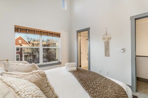 Quarto com uma cama grande, janela com persianas, porta para um banheiro e luminária fixada na parede.