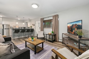 Um apartamento moderno com cozinha, sala de jantar e sala de estar. Possui mesa de centro de madeira, sofá, televisão e uma janela grande com cortinas.