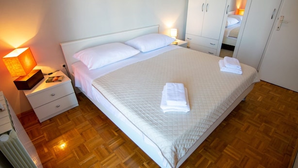 Cama bem arrumada com lençóis e almofadas brancos, mesa de cabeceira com abajur e chão de madeira.