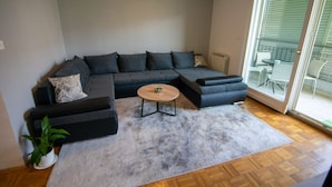 Uma sala de estar com sofá seccional, mesa de centro redonda, tapete felpudo cinza e uma planta em vaso.