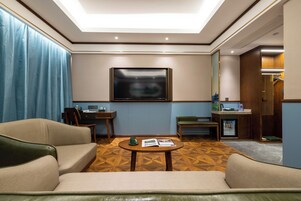 Quarto de hotel com sofá, uma mesa pequena com uma revista, televisão na parede, escrivaninha e mini-frigorífico.