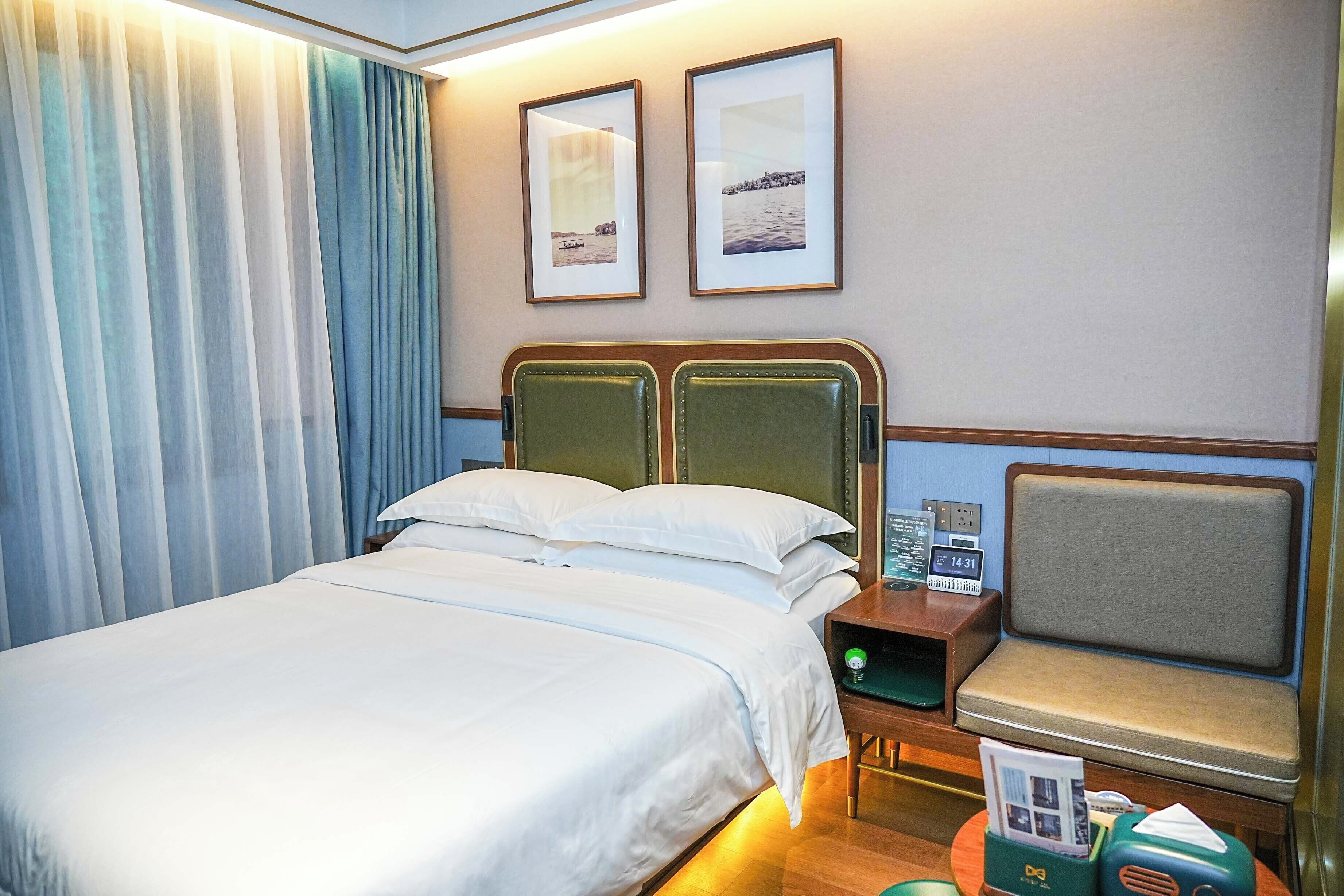 Quarto de hotel com uma cama, mesa de cabeceira com telefone, uma cadeira e duas fotos emolduradas na parede.