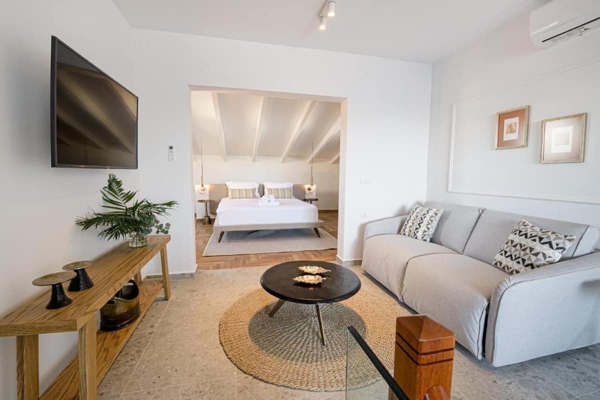 Uma sala de estar moderna com um sofá, uma mesa de centro, uma TV fixada na parede e uma cama ao fundo.