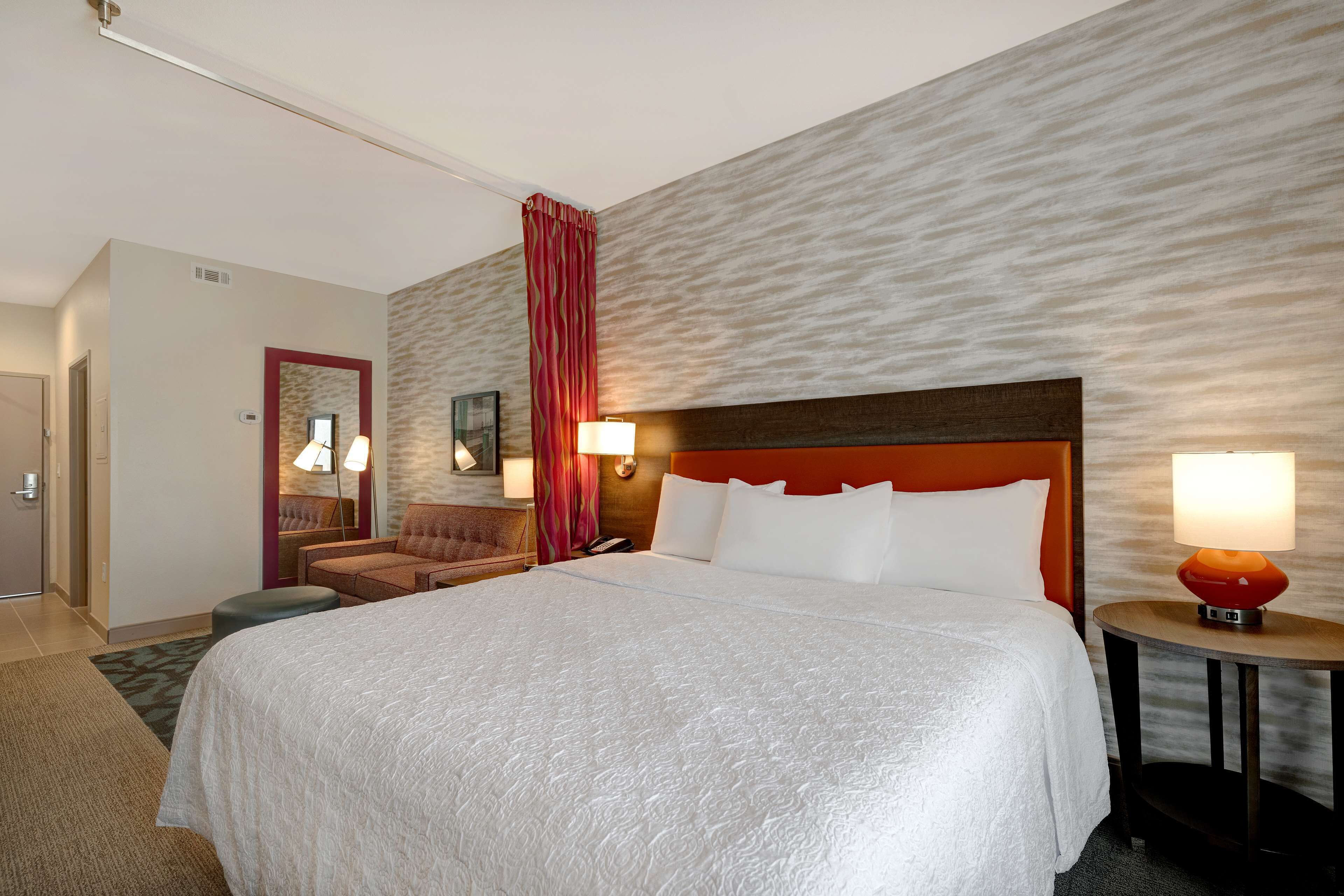Quarto de hotel com uma cama grande, cabeceira de madeira, mesa lateral redonda com abajur, uma cadeira, um pequeno banquinho e um espelho na parede.