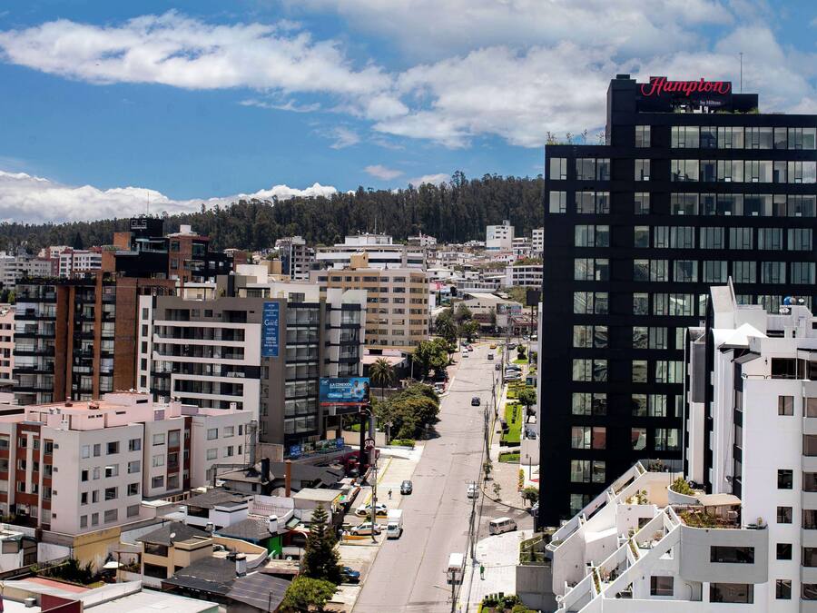 Um hotel moderno com uma placa de destaque "Hampton by Hilton", rodeado por edifícios urbanos e um céu claro.