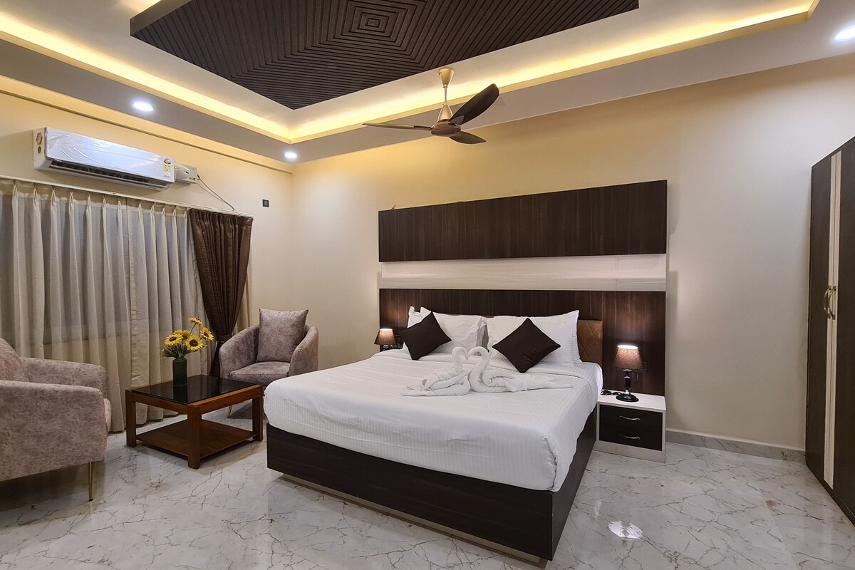 Quarto de hotel moderno com uma cama grande, duas poltronas, uma mesinha de centro com um vaso de flores, um ventilador de teto, um aparelho de ar condicionado e um guarda-roupa.