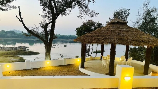 Um gazebo com telhado de palha, cadeiras brancas e luzes iluminadas, cercado por uma parede branca e um caminho de terra.