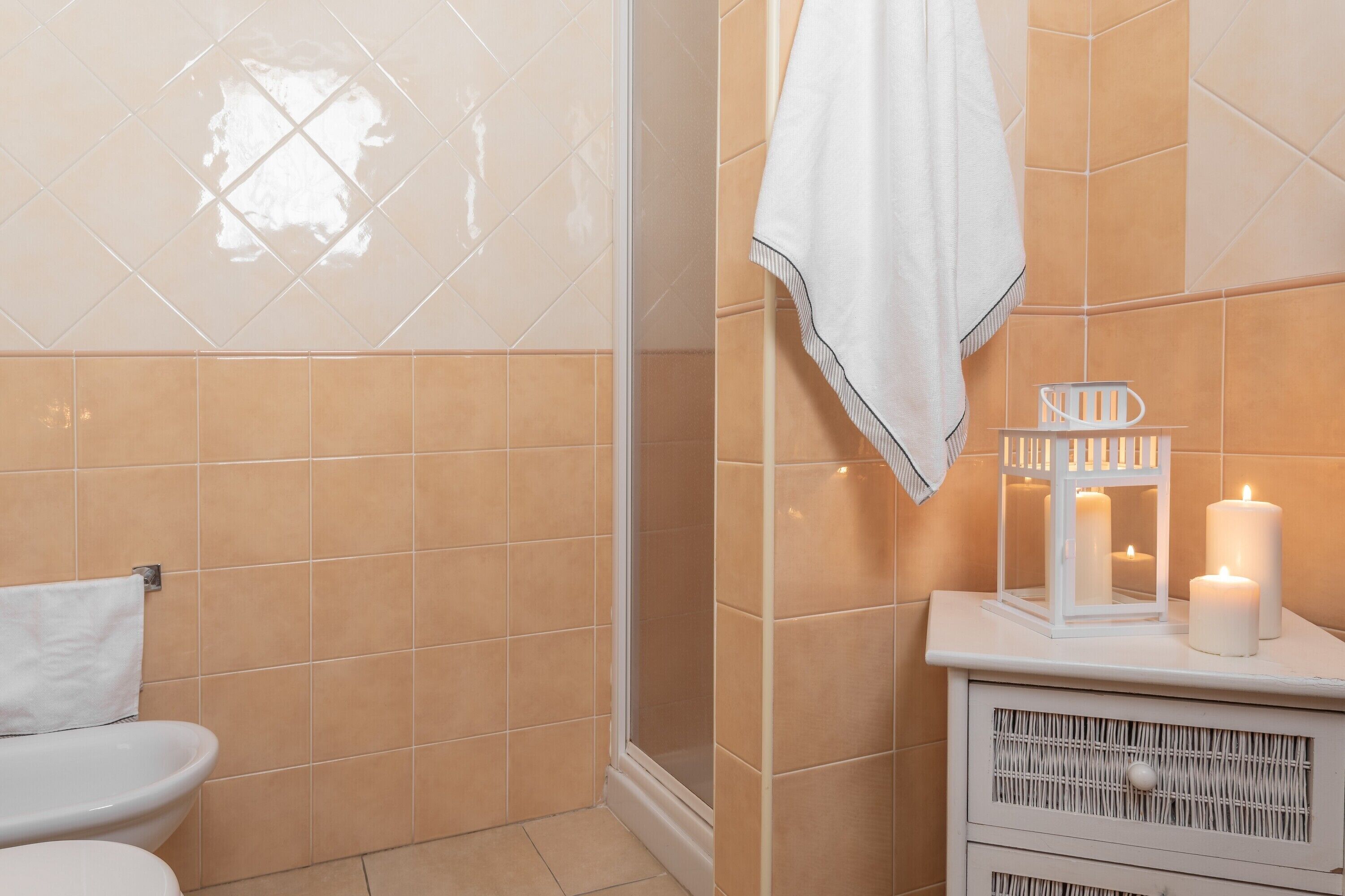 Casa de banho com azulejos bege, uma toalha branca pendurada na parede, sanita branca e toucador com velas.
