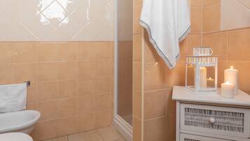 Casa de banho com azulejos bege, uma toalha branca pendurada na parede, sanita branca e toucador com velas.