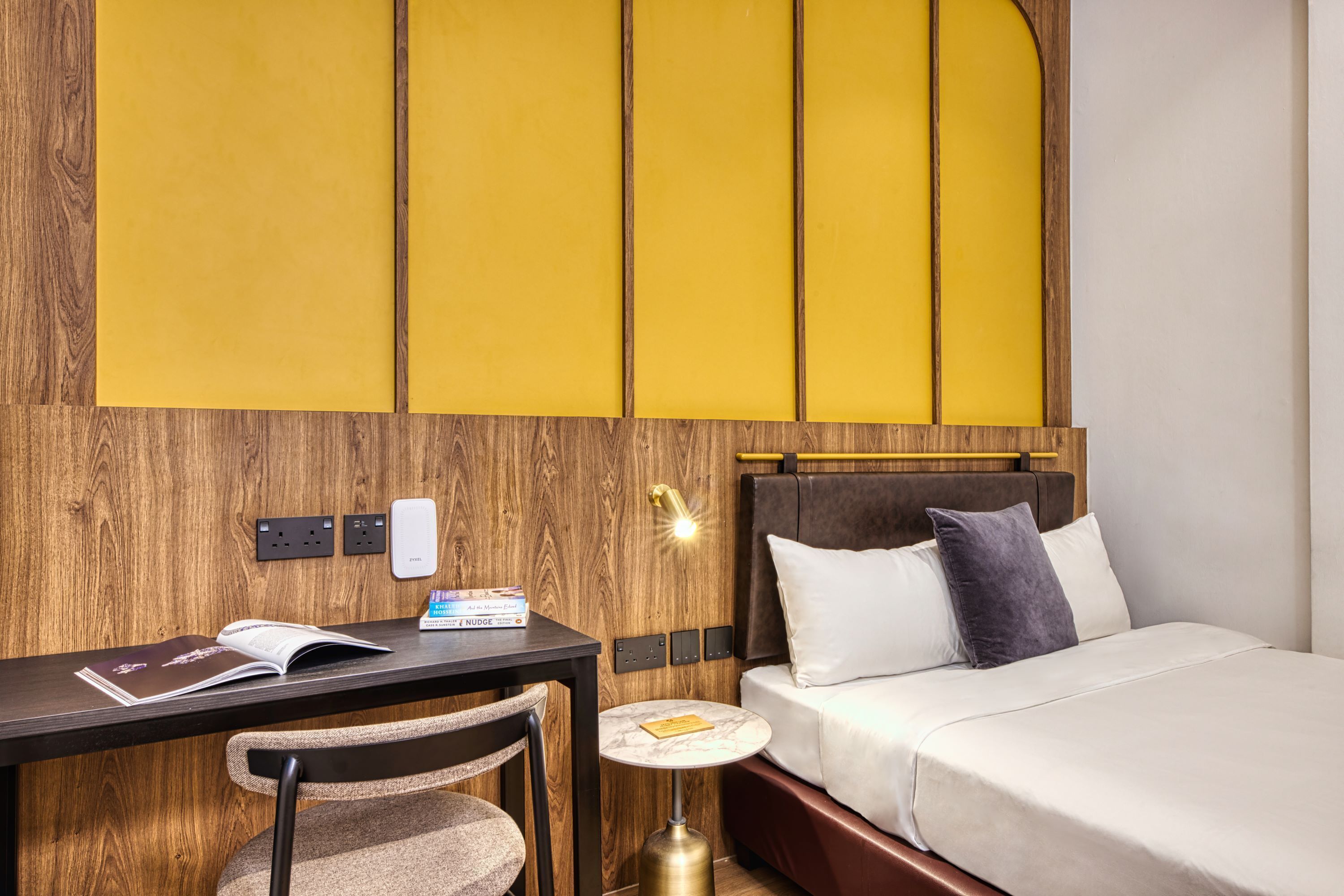 Quarto de hotel com cabeceira de madeira, cama com almofadas brancas e cinzentas, uma pequena mesa redonda com um livro, uma cadeira e uma mesinha de cabeceira com um abajur.