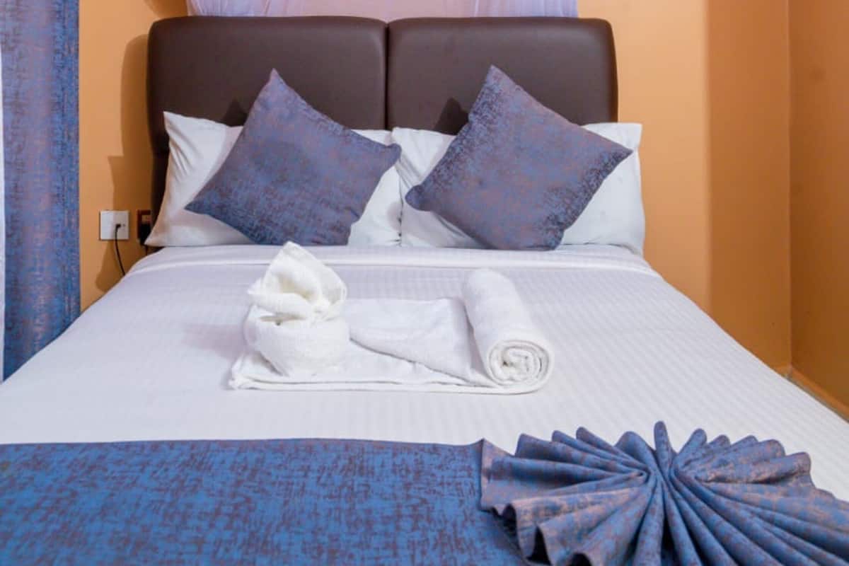 Cama bem arrumada com colcha branca e azul, dois travesseiros e uma toalha branca enrolada.