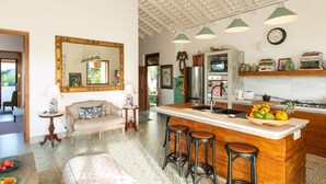 Uma sala de estar ampla com sofá, mesa de centro e um espelho grande. Há uma cozinha com ilha de madeira, banquetas e uma fruteira.