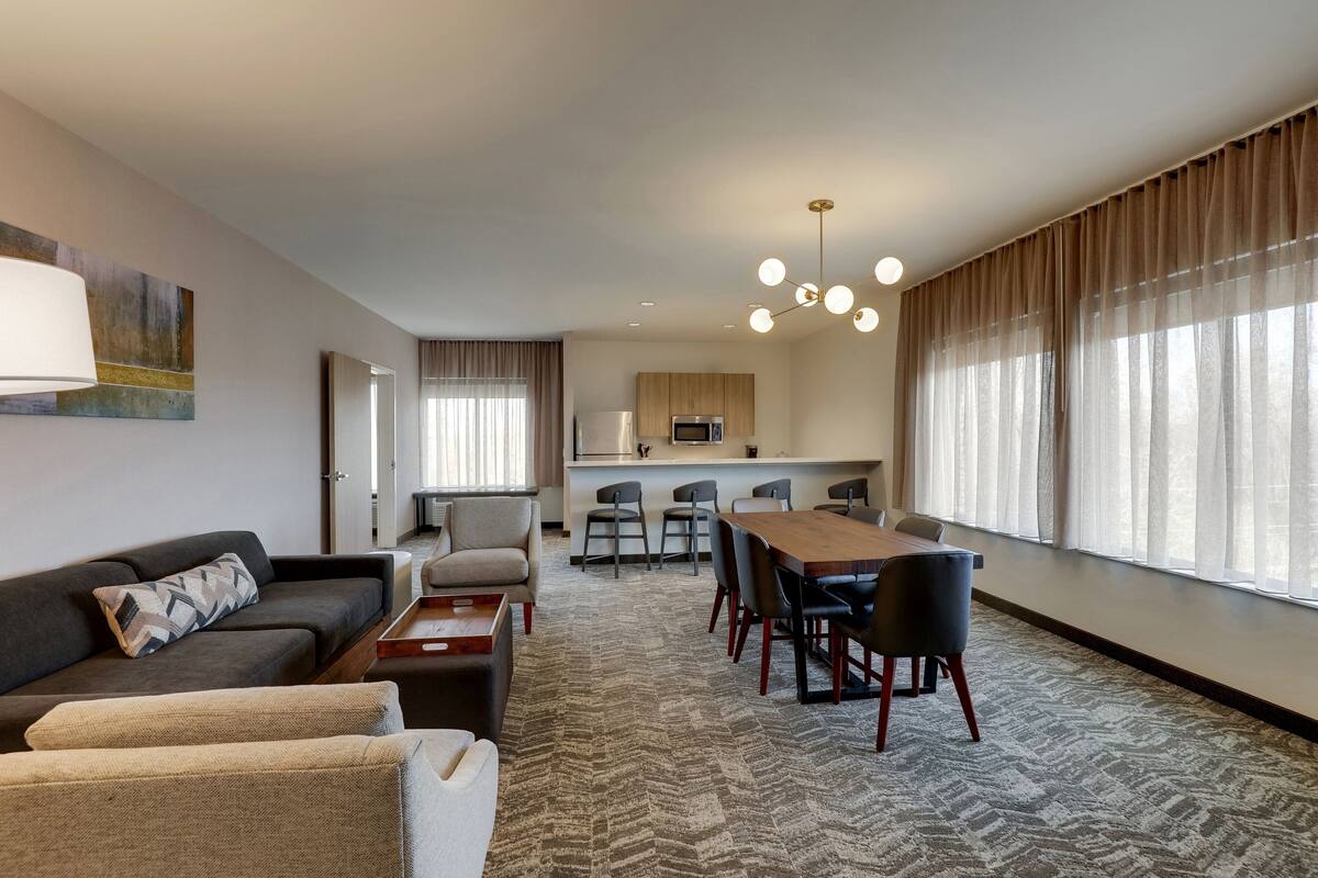 Quarto de hotel moderno com sofá, poltronas, mesa de jantar com cadeiras, área de cozinha e janelas amplas com cortinas.