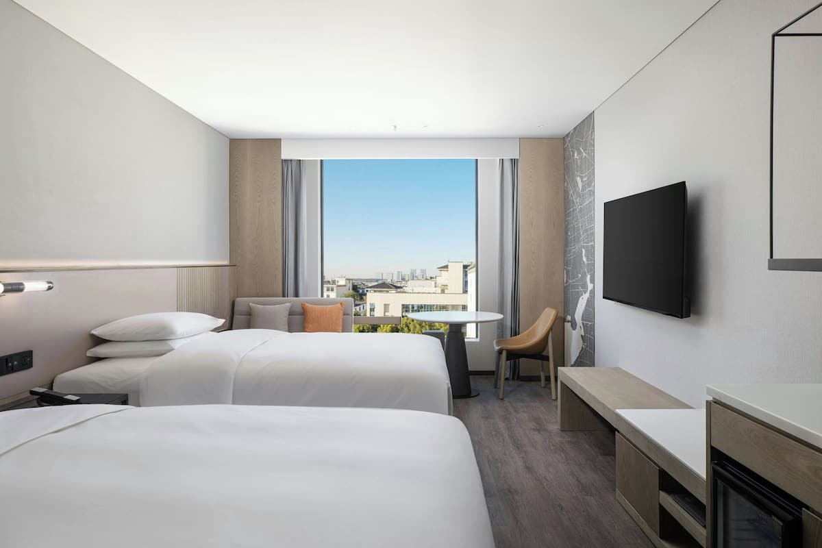 Quarto de hotel com duas camas, uma mesa, uma cadeira, uma televisão e janela com vista para a cidade.
