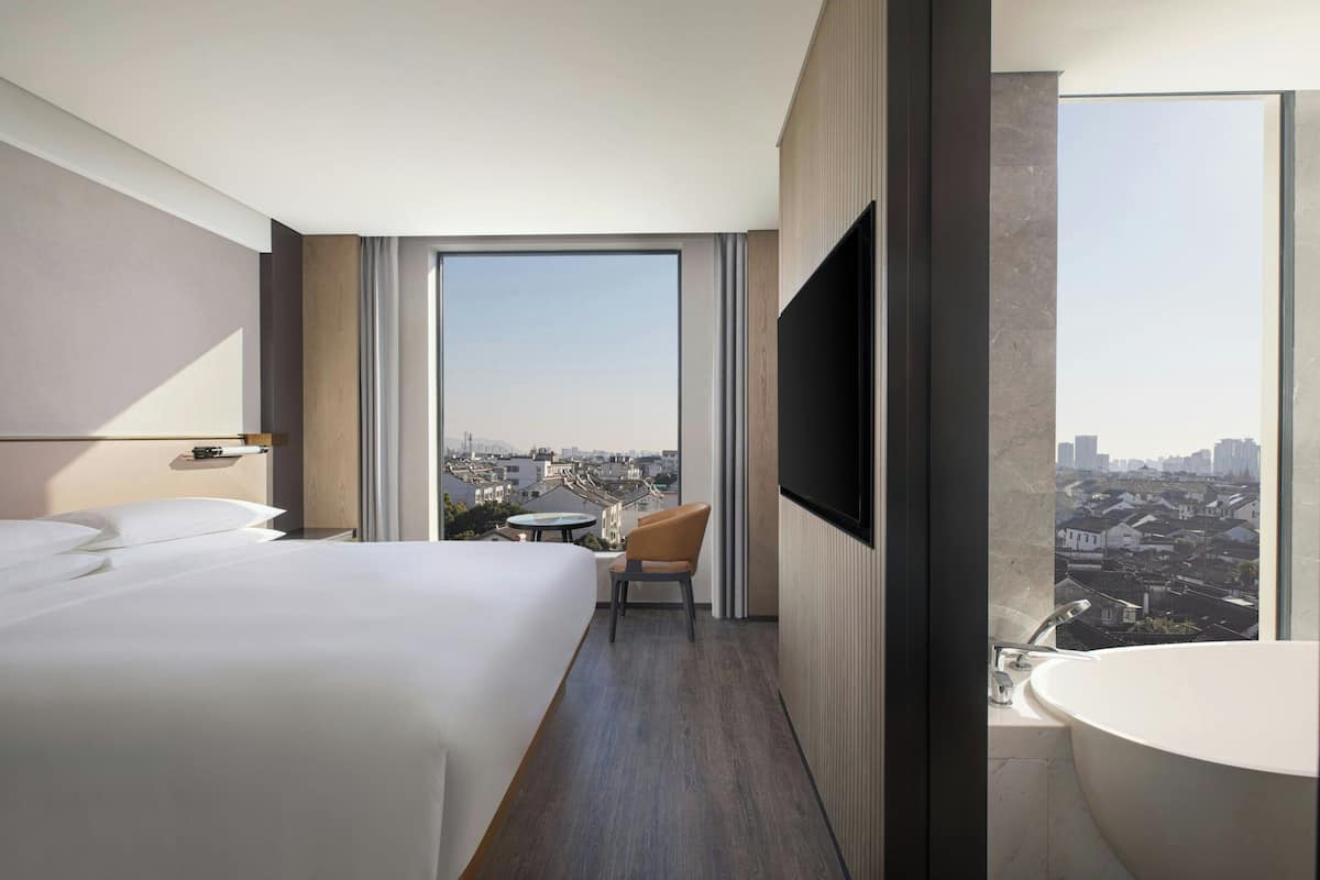 Quarto de hotel moderno com uma cama grande, televisão, banheiro com banheira e vista para a cidade.