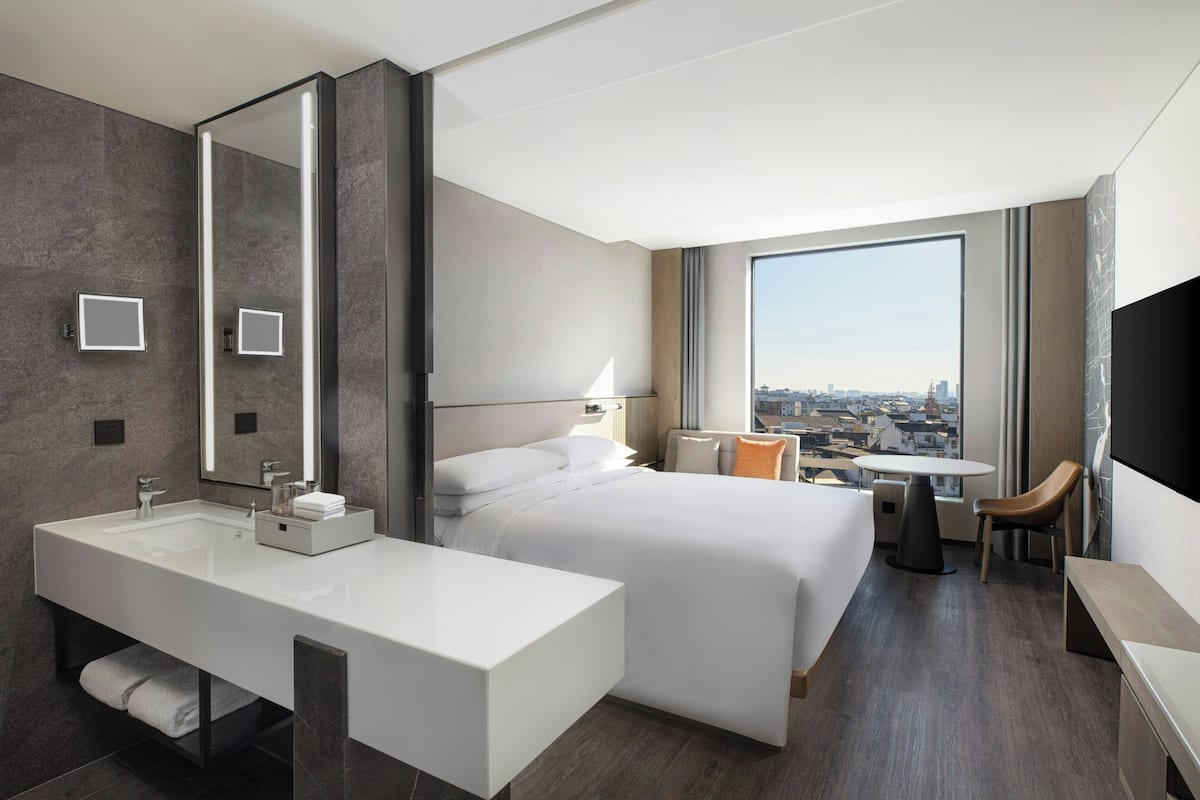 Quarto de hotel moderno com uma cama grande, área de estar separada, escrivaninha, janela ampla com vista para a cidade e banheiro com um espelho grande e pia.