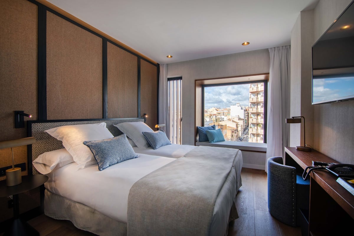 Quarto de hotel com duas camas, uma escrivaninha, uma cadeira, televisão e uma janela grande com vista para a cidade.