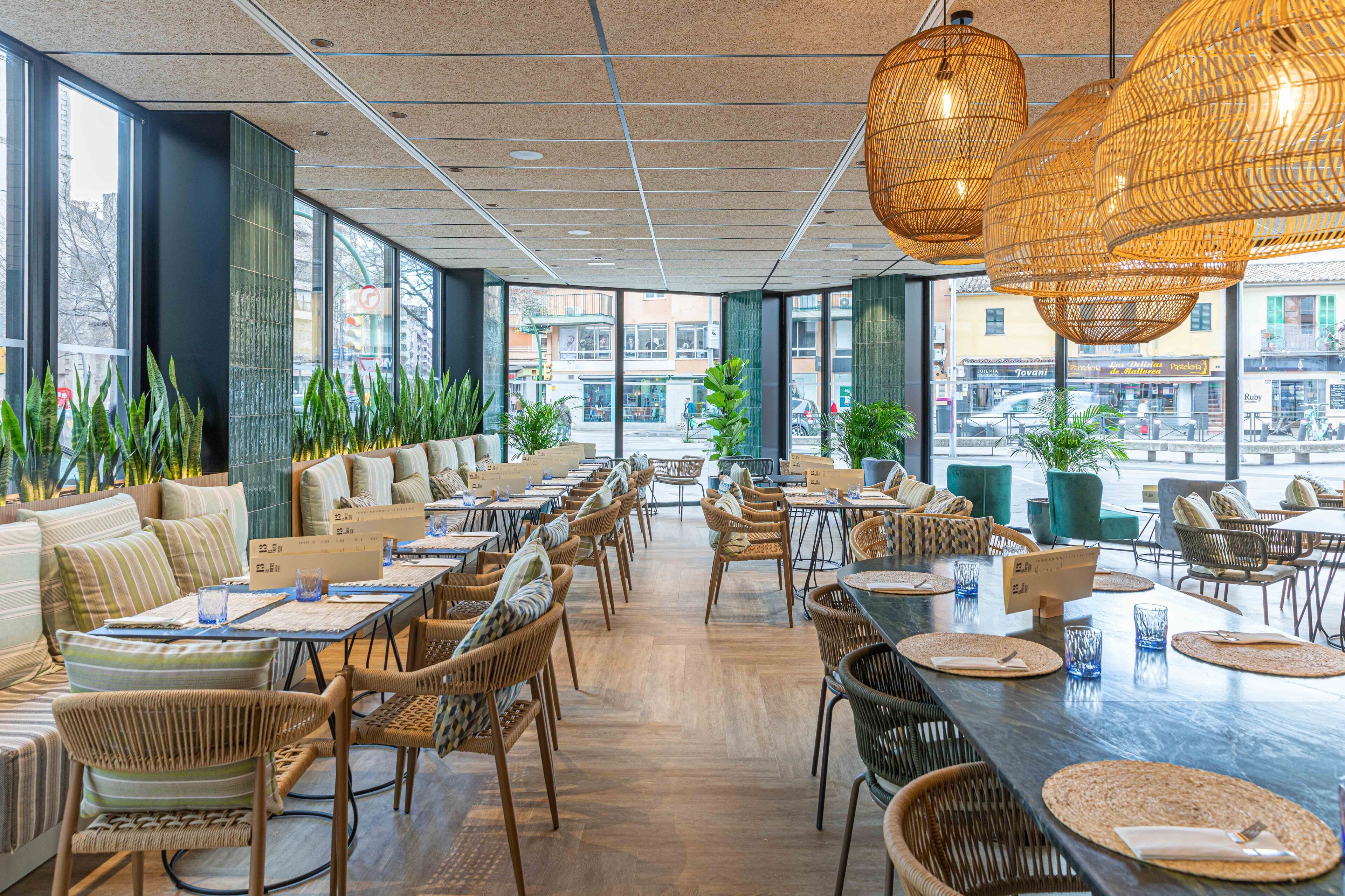 Um restaurante moderno com mesas comunitárias grandes, cadeiras de vime e amplas janelas com vista para a cidade.