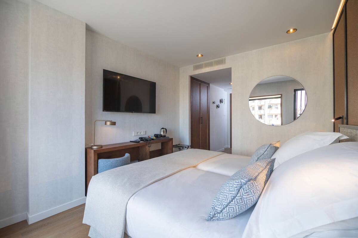 Quarto de hotel com uma cama grande, uma escrivaninha com cadeira, uma televisão de tela plana na parede e um espelho redondo na parede.