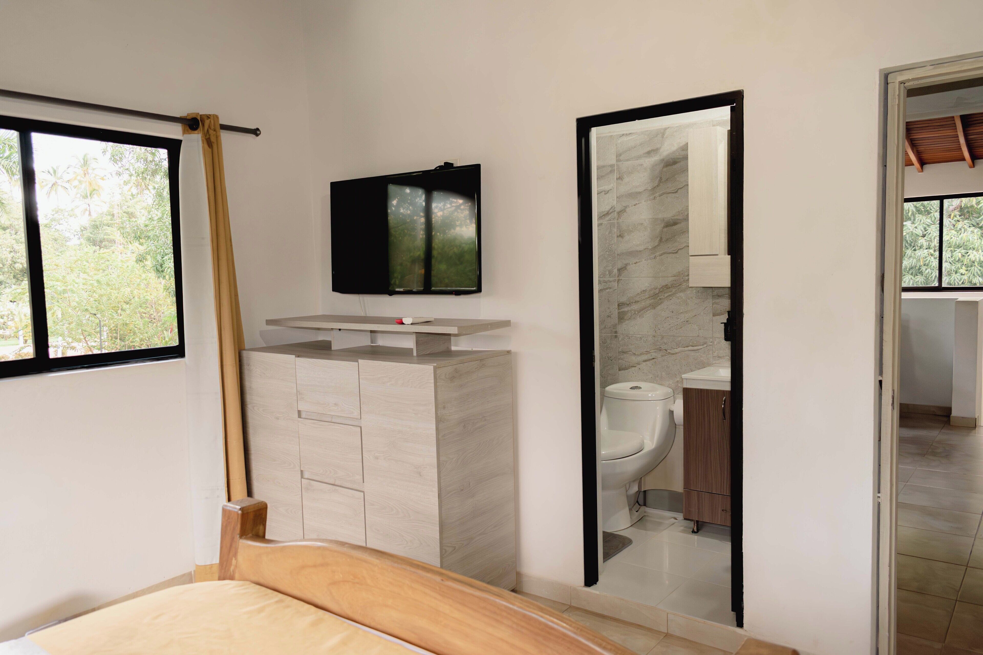 Quarto com cama de madeira, cômoda branca, televisão na parede e banheiro visível através de uma porta aberta.