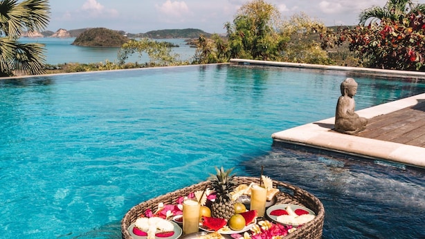 Uma área de piscina tranquila com uma cesta flutuante contendo comida e bebidas, rodeada por vegetação exuberante e vista clara para o mar.