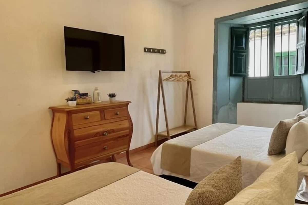 Quarto de hotel com duas camas, cômoda de madeira, ventilador de teto, televisão na parede e janela com persianas.