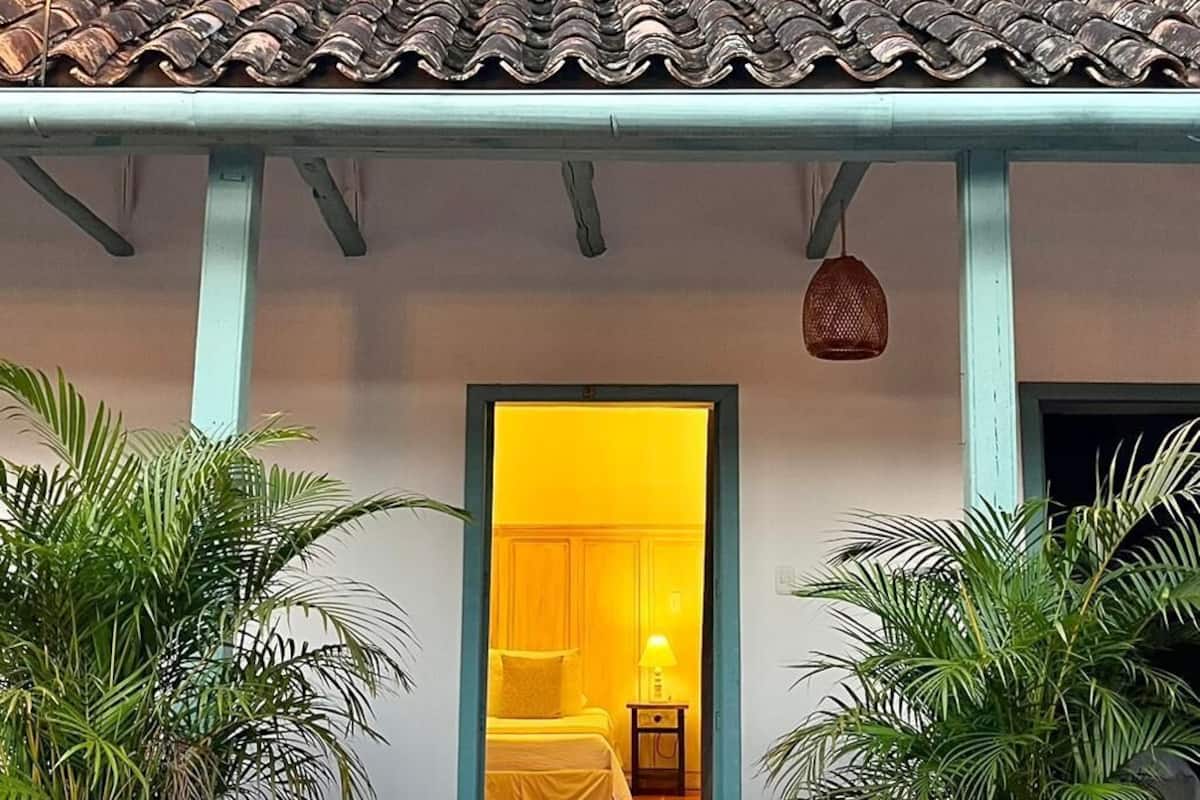 Quarto de hotel com telhado de azulejos, uma cama, mesa de cabeceira e abajur. Existem duas poltronas com almofadas na parte externa do quarto.