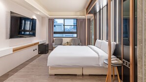 Quarto de hotel moderno com uma cama grande, uma televisão presa na parede, uma mesinha com um telefone e uma janela com cortinas.
