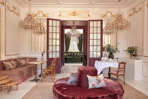Um quarto luxuoso com um sofá de veludo vermelho, um banquinho redondo, uma mesa de jantar com cadeiras e um lustre.