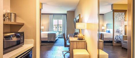 Quarto de hotel com cama, escrivaninha com luminária, cadeira, micro-ondas e um espelho grande.