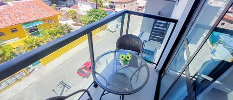 Varanda com mesa de vidro e duas cadeiras, vista para uma rua com edifícios e céu claro.