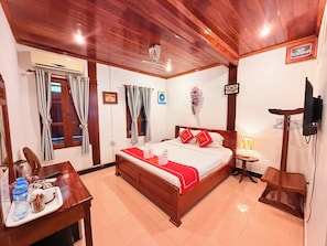 Quarto de hotel com teto de madeira, cama com roupa de cama vermelha e branca, mesa de cabeceira com abajur e TV na parede.