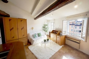 Um espaço compacto com sofá branco, mesa redonda pequena e tapete felpudo branco. Inclui um guarda-roupa de madeira, radiador e janela com vista para edifícios.
